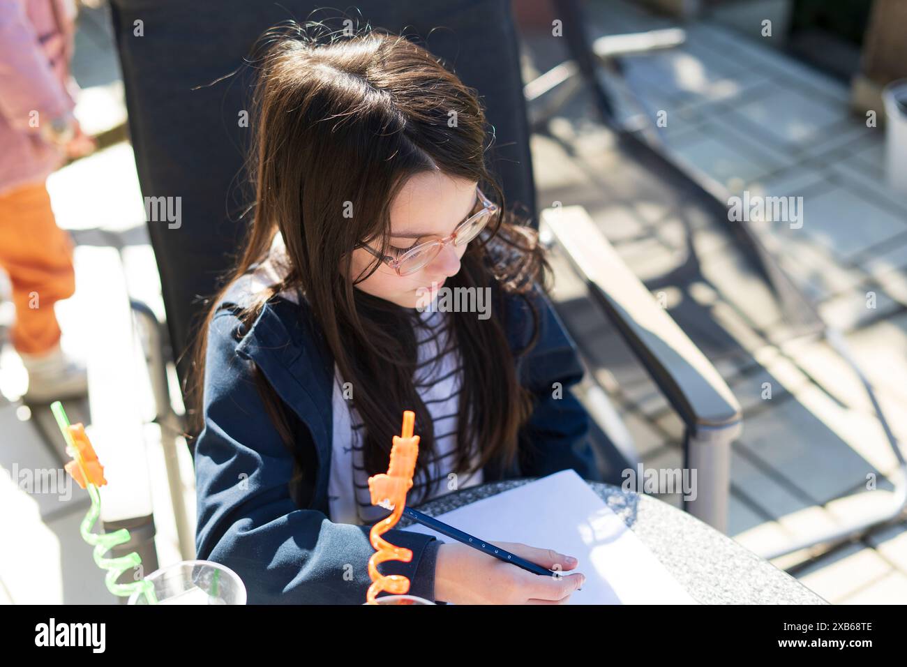 A little girl with glasses sits at a table in the backyard and does her ...