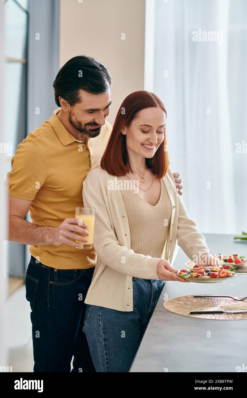A redhead woman and bearded man laugh and eat meal in a modern kitchen ...