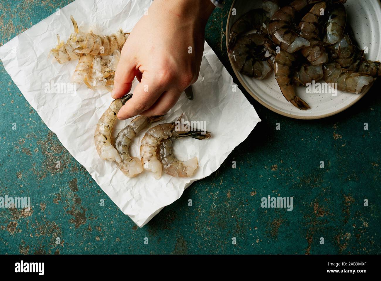 Hands arranging peeled fresh shrimp on a rustic surface with a plate of ...