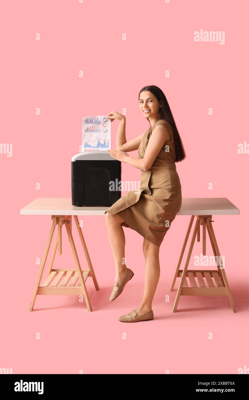 Young woman destroying documents using shredder on table against pink ...