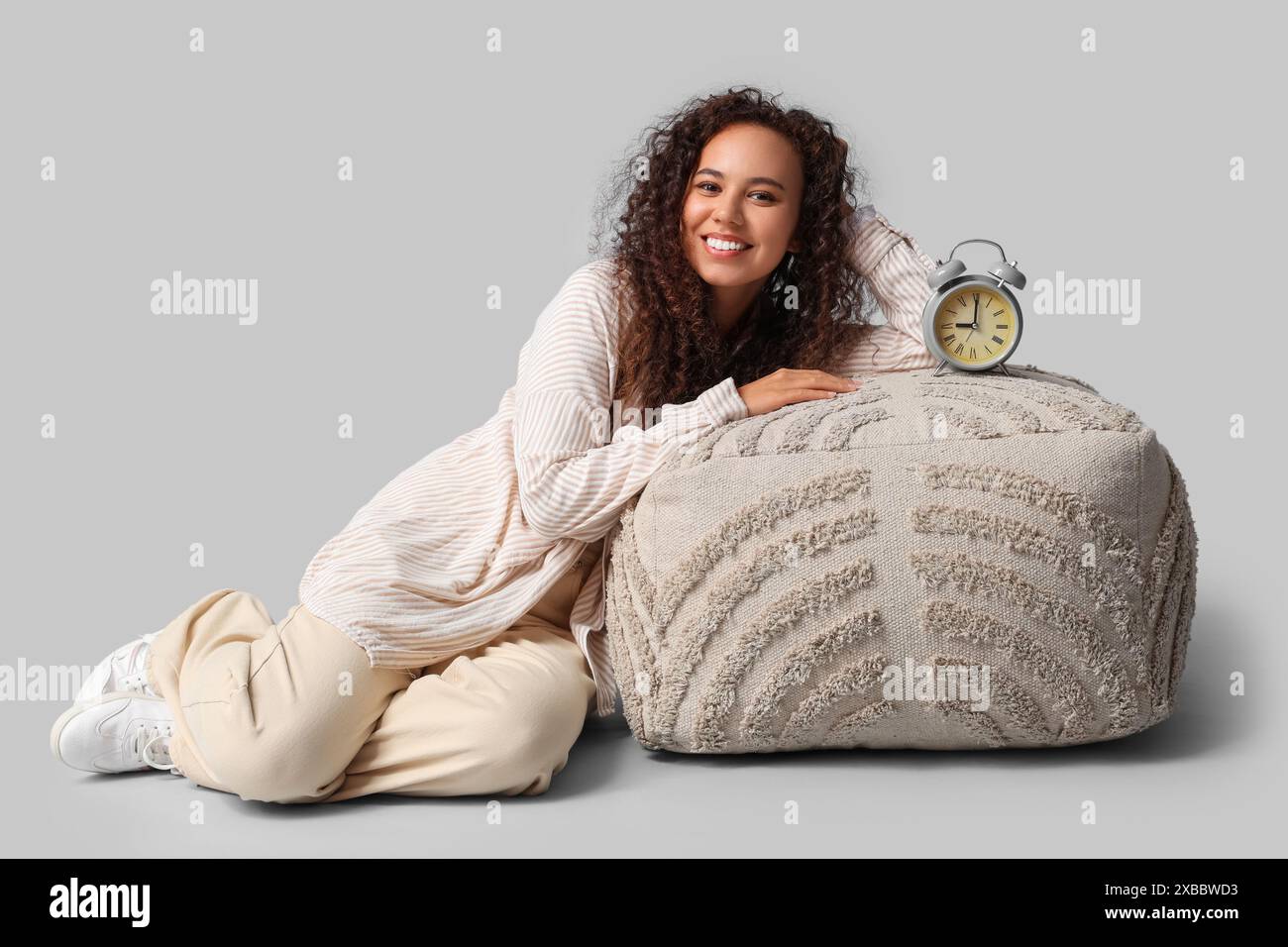 Beautiful young African-American woman with alarm clock resting on pouf ...