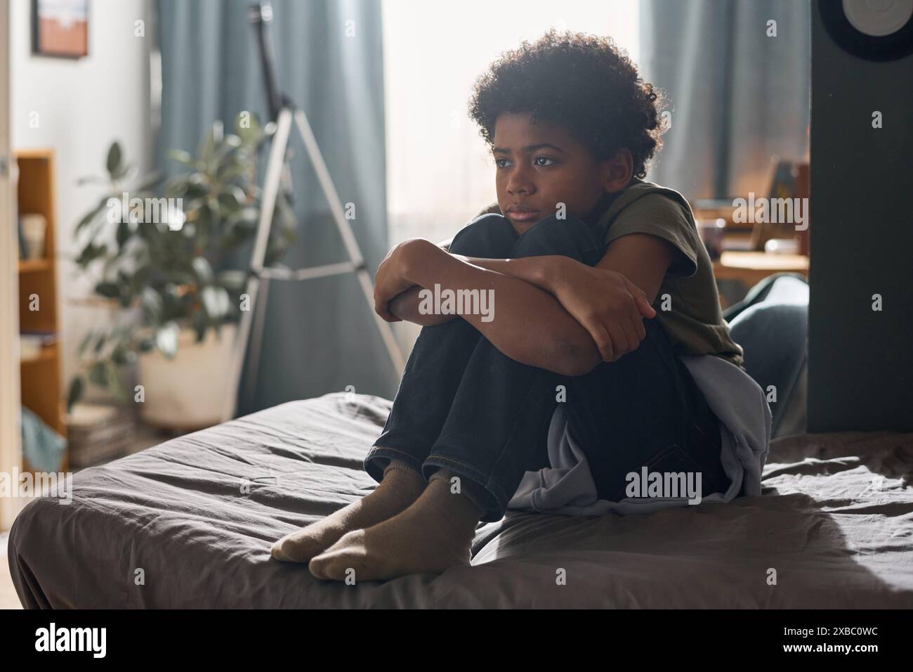 Full length portrait of upset young African American boy sitting on bed ...