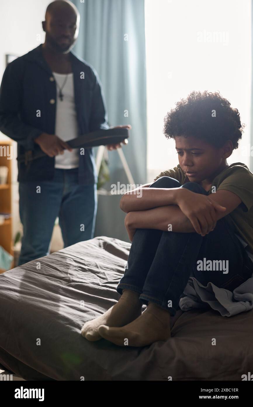 Vertical full length portrait of young Black boy crying sitting on bed ...