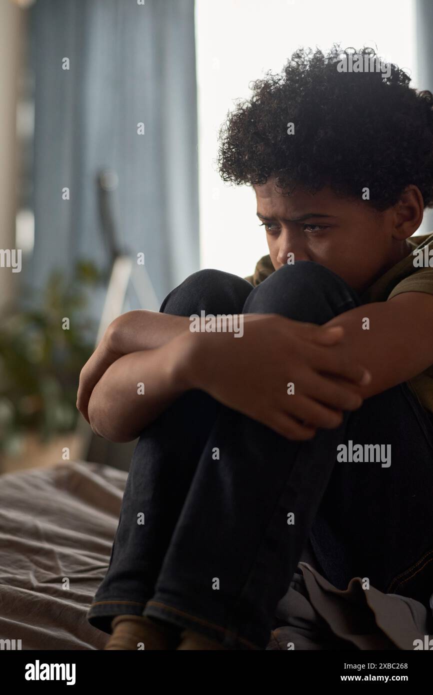 Vertical portrait of crying young boy sitting on bed in his room and ...