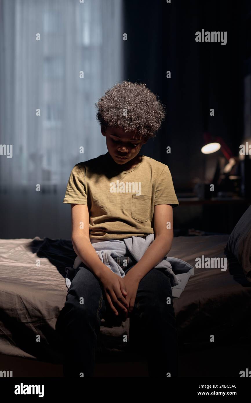 Vertical portrait of young Black boy sitting on bed with dark dramatic ...