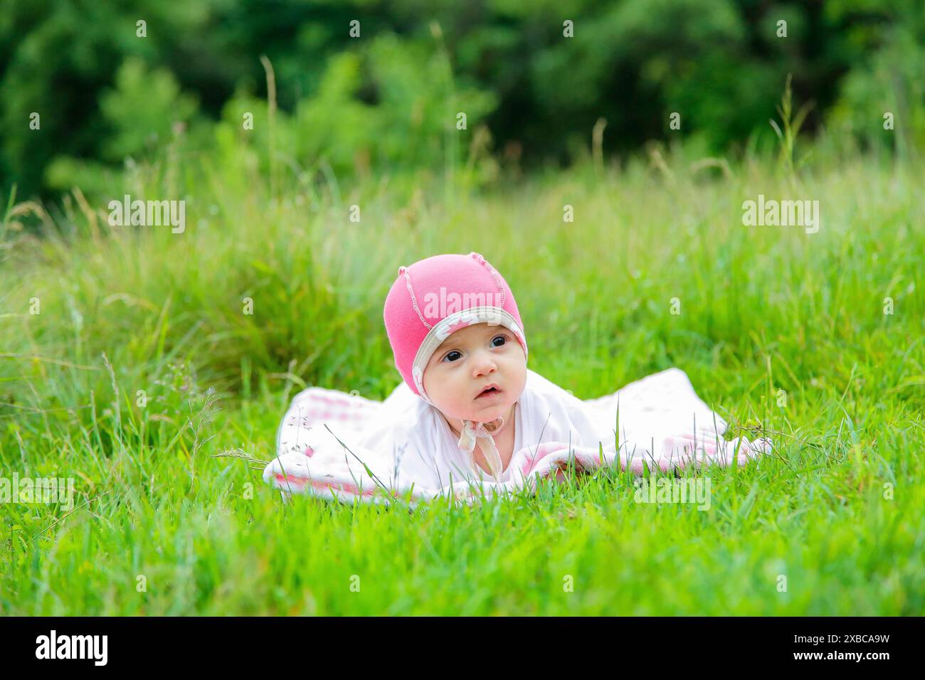 Baby lying on a white blanket in the grass, wearing a pink hat, surrounded by green foliage ...