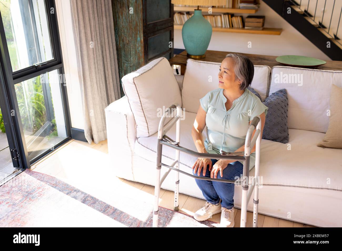 Asian senior woman using walker, sitting on couch in bright living room ...
