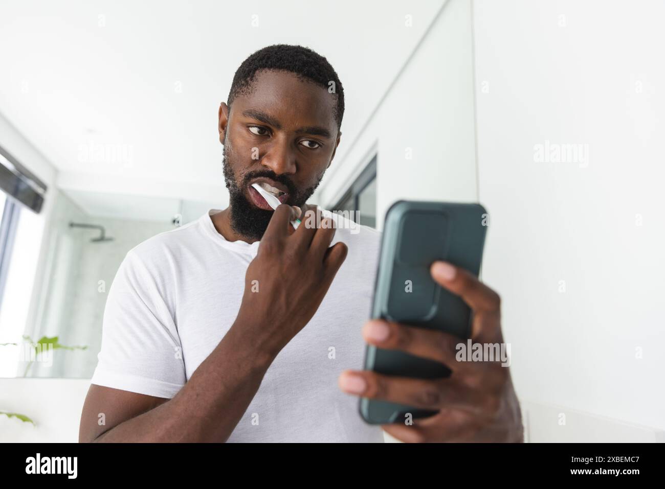 African American young man brushing teeth while looking at smartphone ...