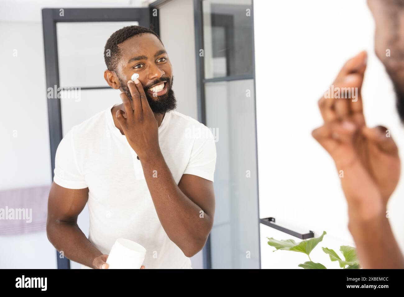 African American young man applying face cream while smiling at his ...