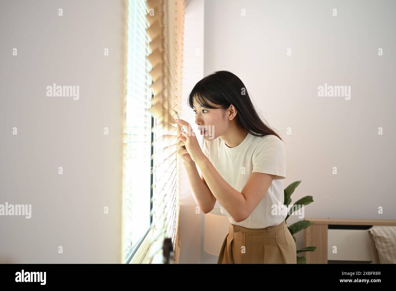 Young Asian woman looking outside through Venetian window blinds at ...