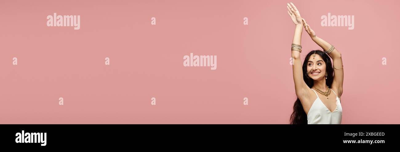 A young indian woman enthusiastically raises her arms on a vibrant pink ...