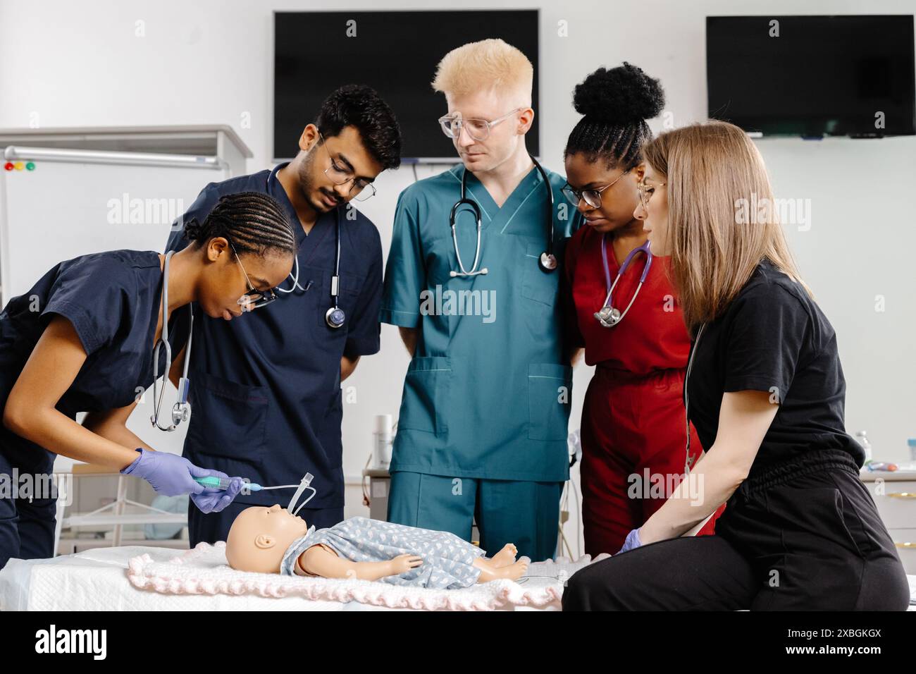 Four medical students in scrubs are gathered around a training dummy to ...