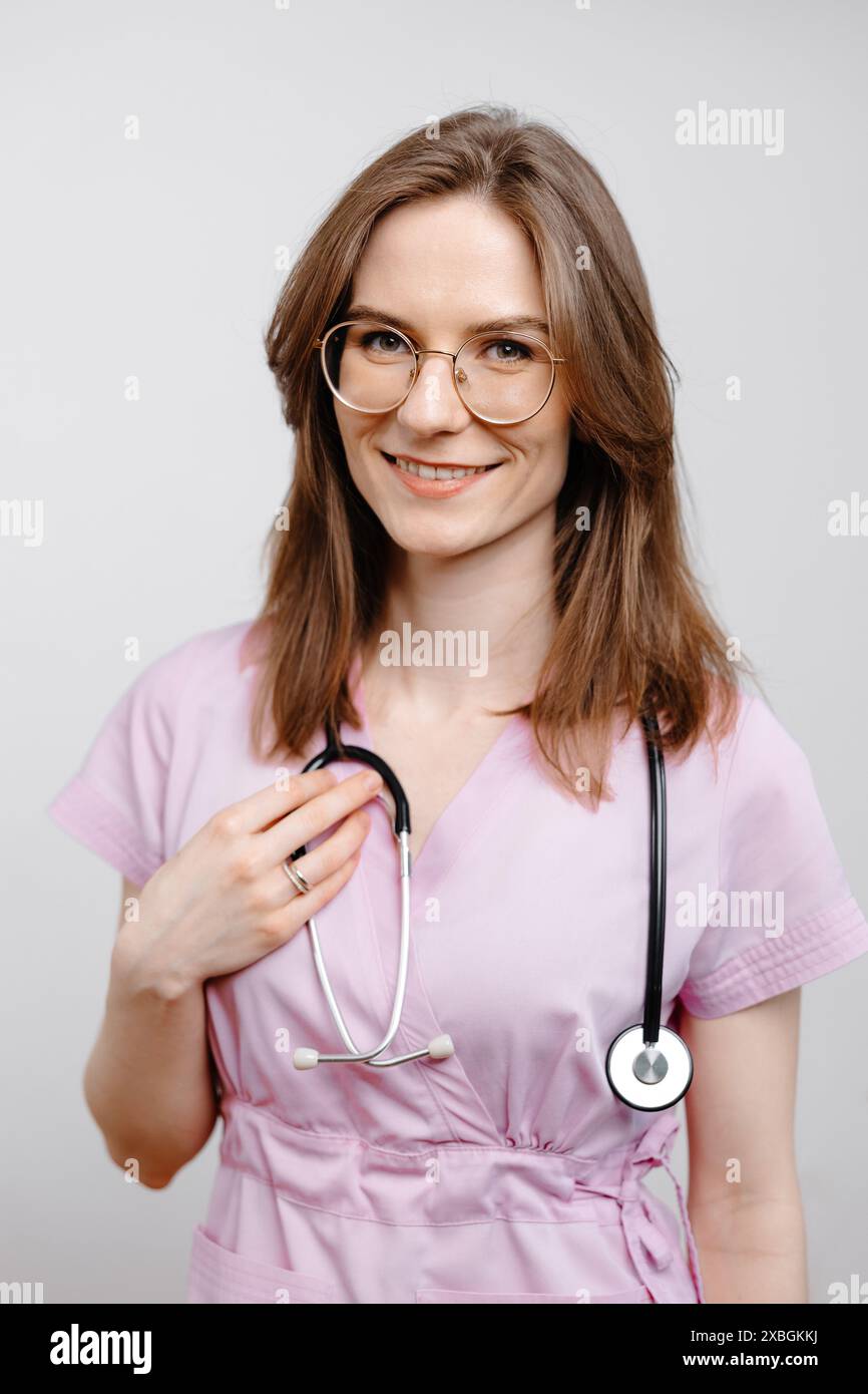 A smiling female doctor in pink scrubs holds a stethoscope against her ...