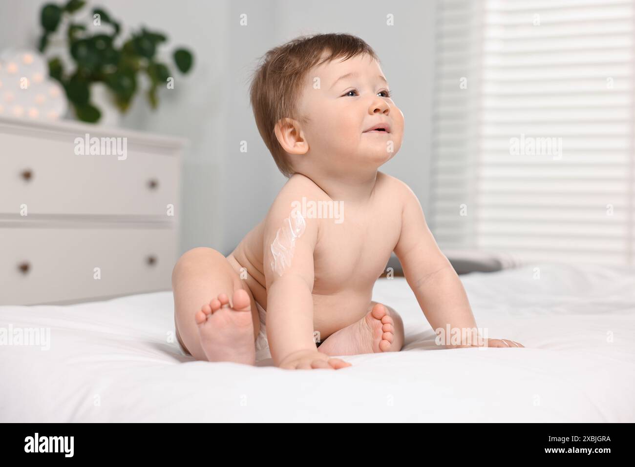 Cute little baby with moisturizing cream onto body sitting on bed at ...
