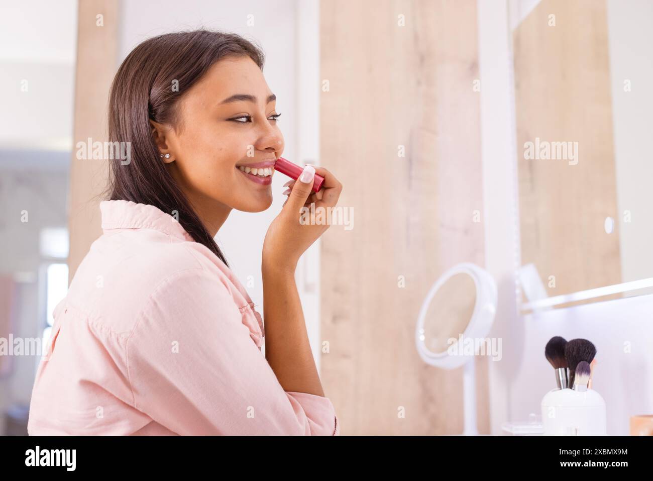 Happy biracial woman applying lipstick, smiling at her reflection in ...