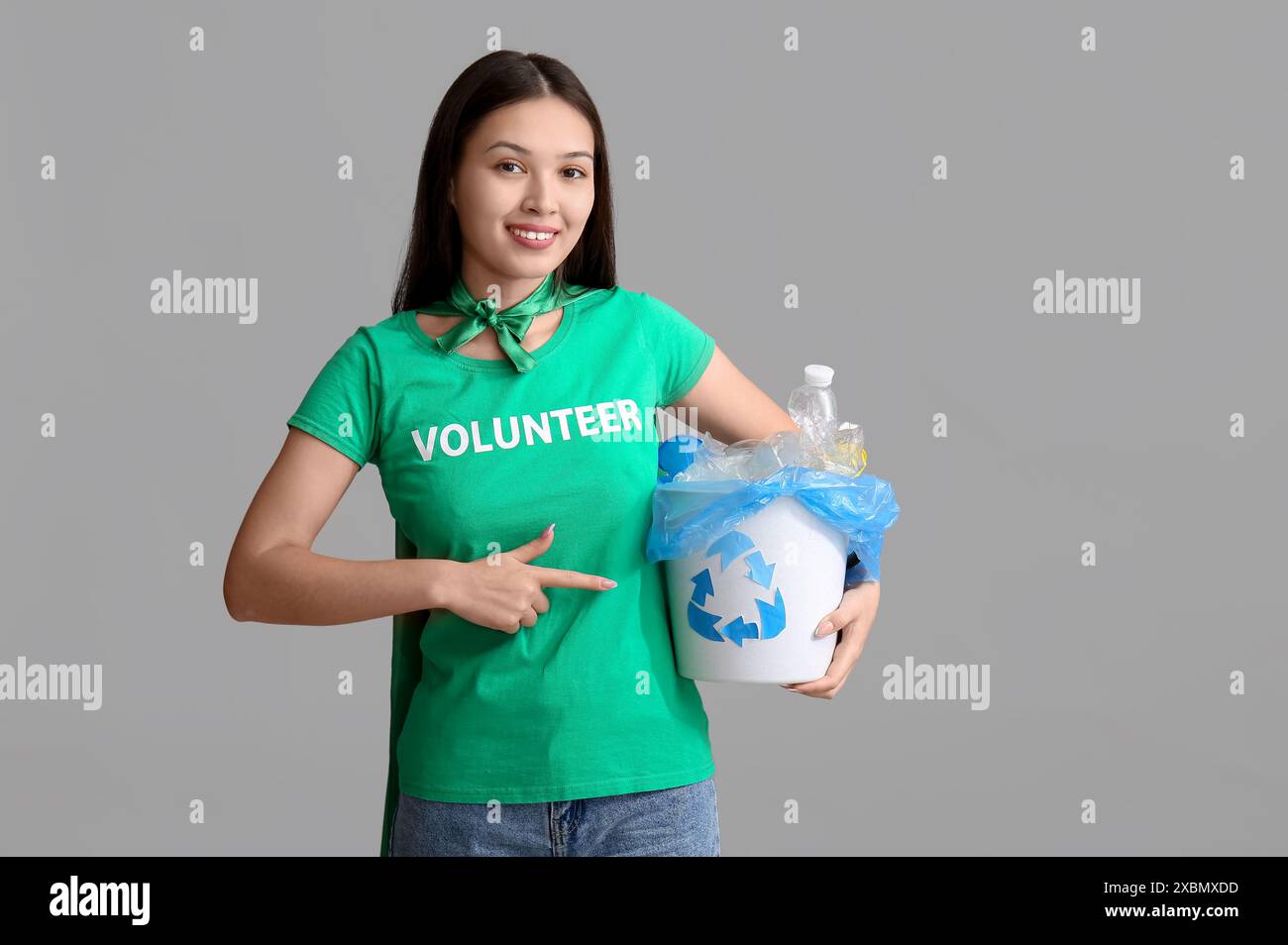 Young female volunteer in superhero cape pointing at recycle bin with garbage on white ...