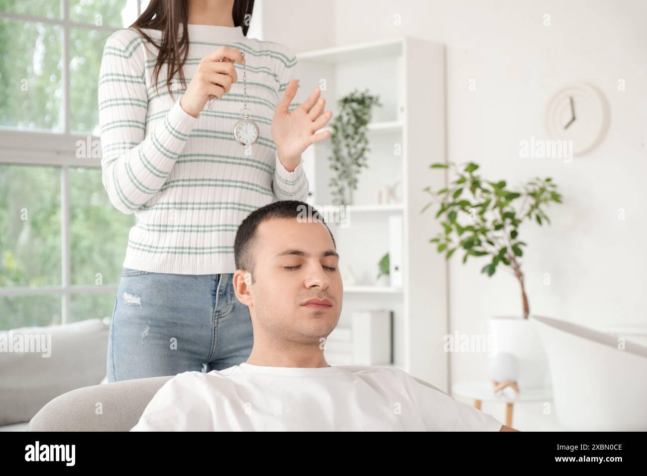 Female psychologist with pocket watch hypnotizing young man on armchair ...