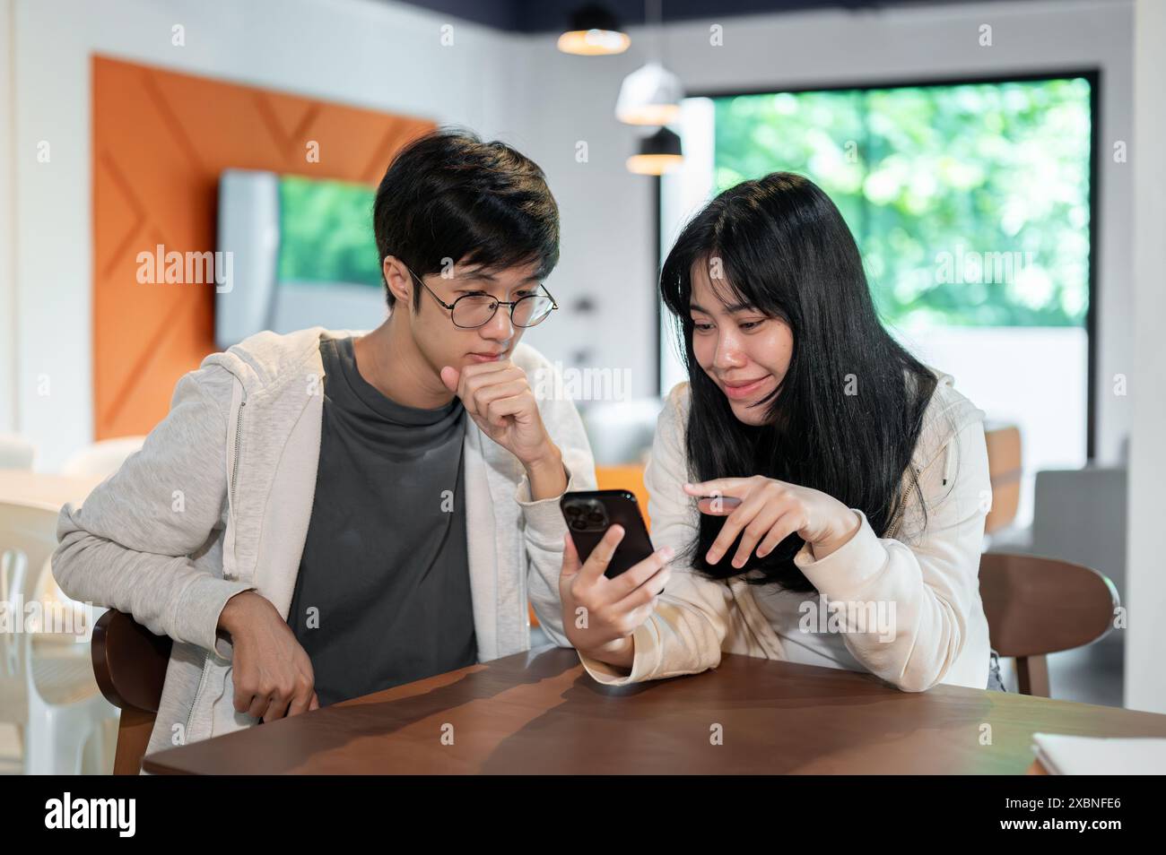 Two young Asian friends are looking at a smartphone screen and talking ...