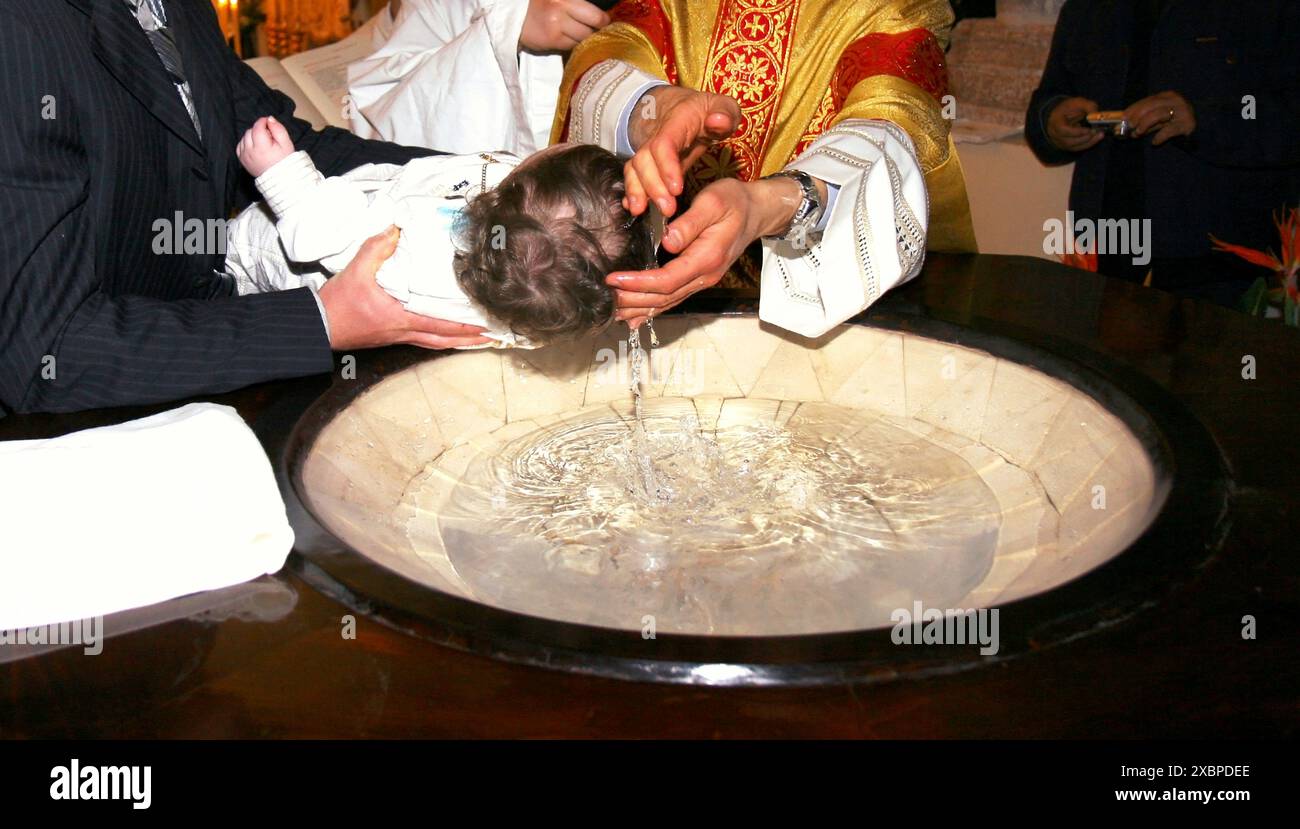 Priest wearing ceremonial clothing pouring water on baby's head held by ...