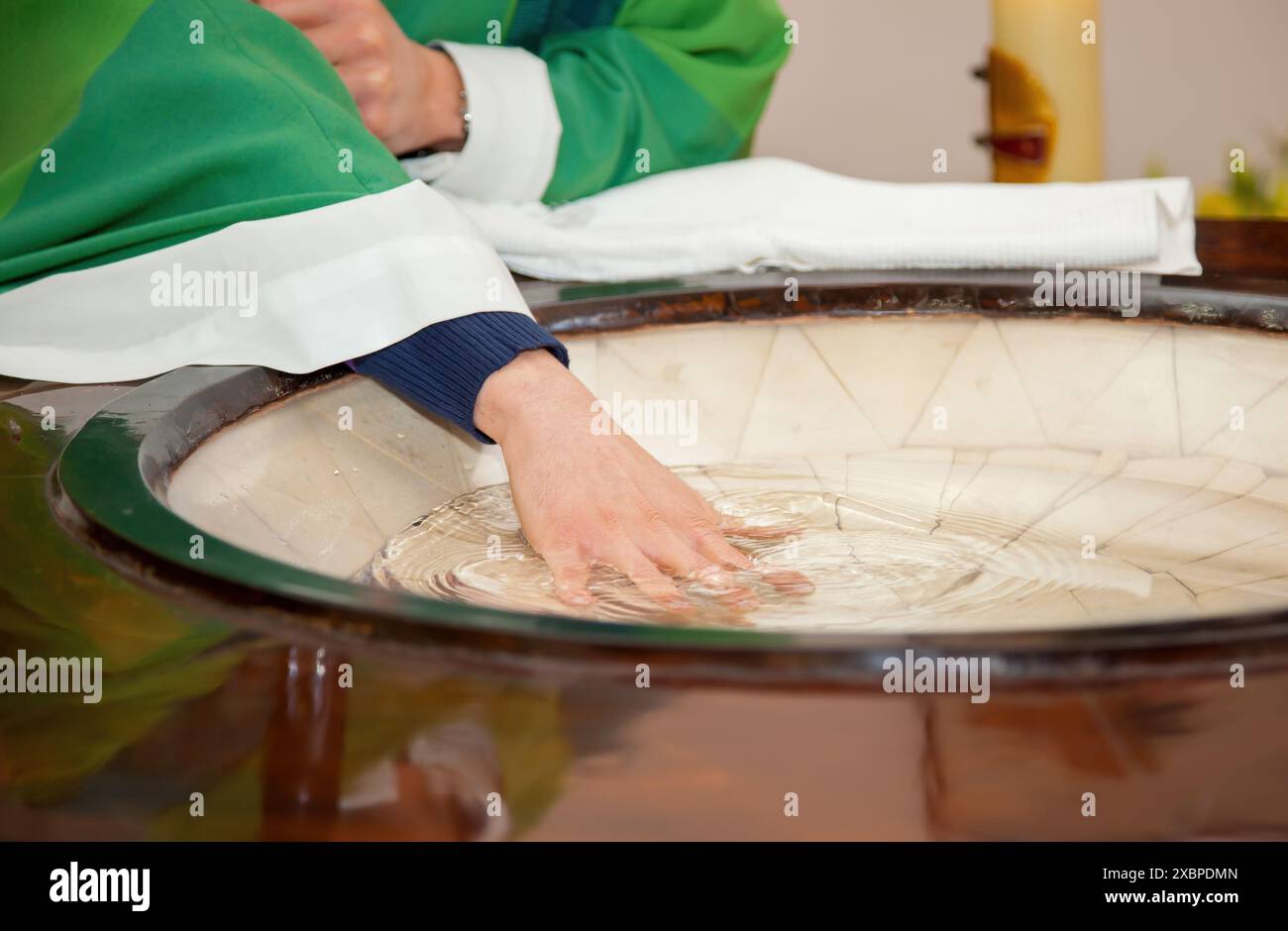 Priest wearing green liturgical vestments touching holy water inside a marble baptismal font in ...