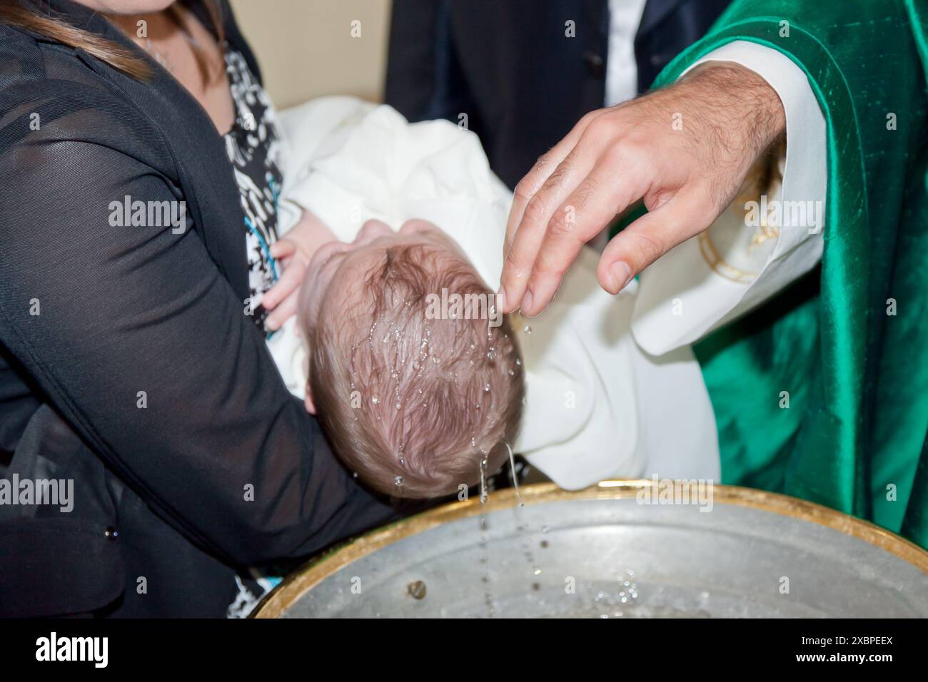 Priest wearing green chasuble pouring water on baby's head held by his ...