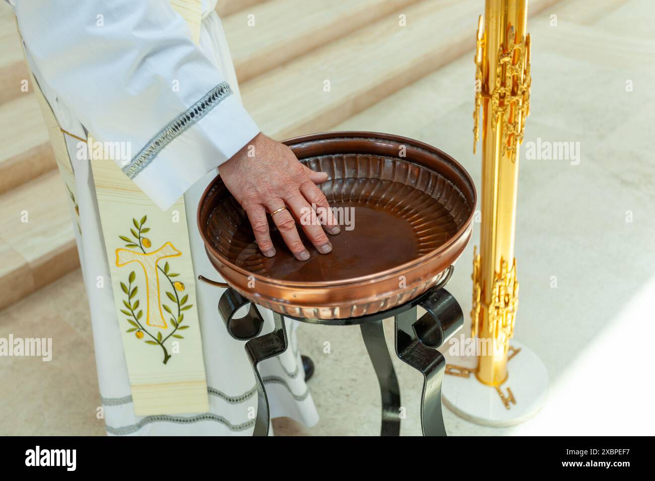 Priest wearing a white clergy shirt and stole is blessing the water of a copper baptismal font ...