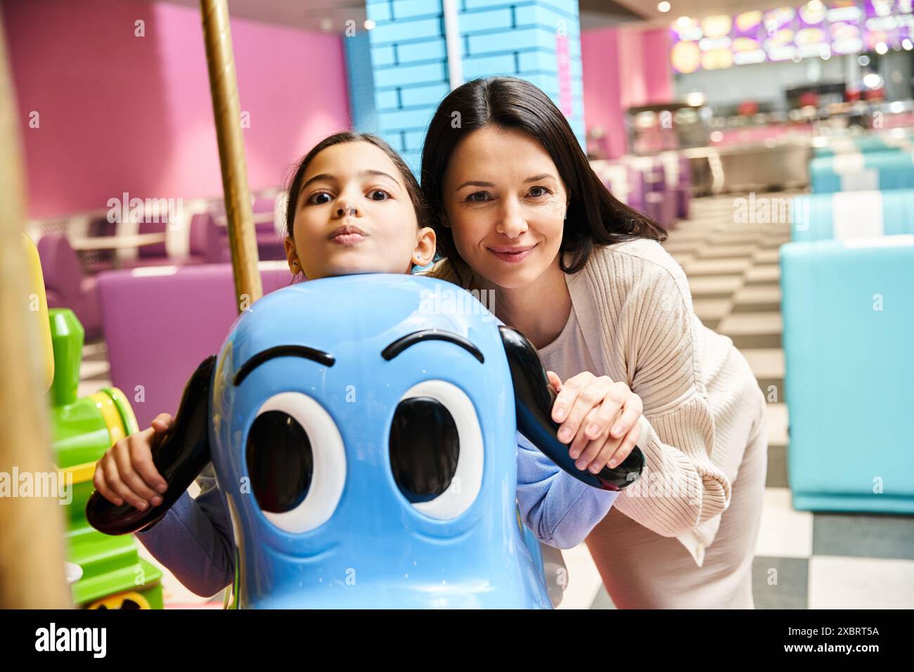 A cheerful woman and child joyfully posing together next to a colorful carousel toy in a mall ...