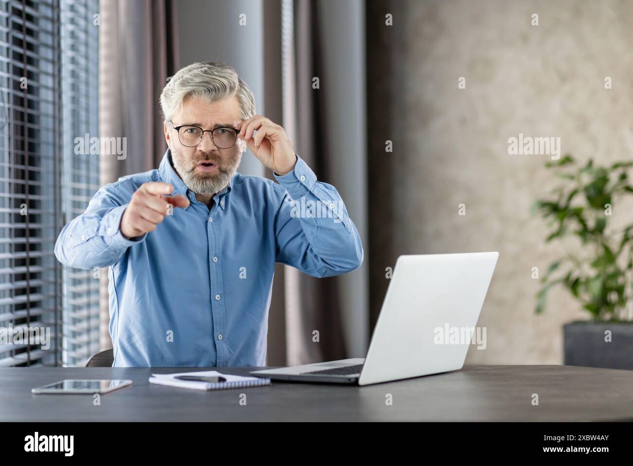 Elderly man in blue shirt pointing at camera, working remotely from home office with laptop ...
