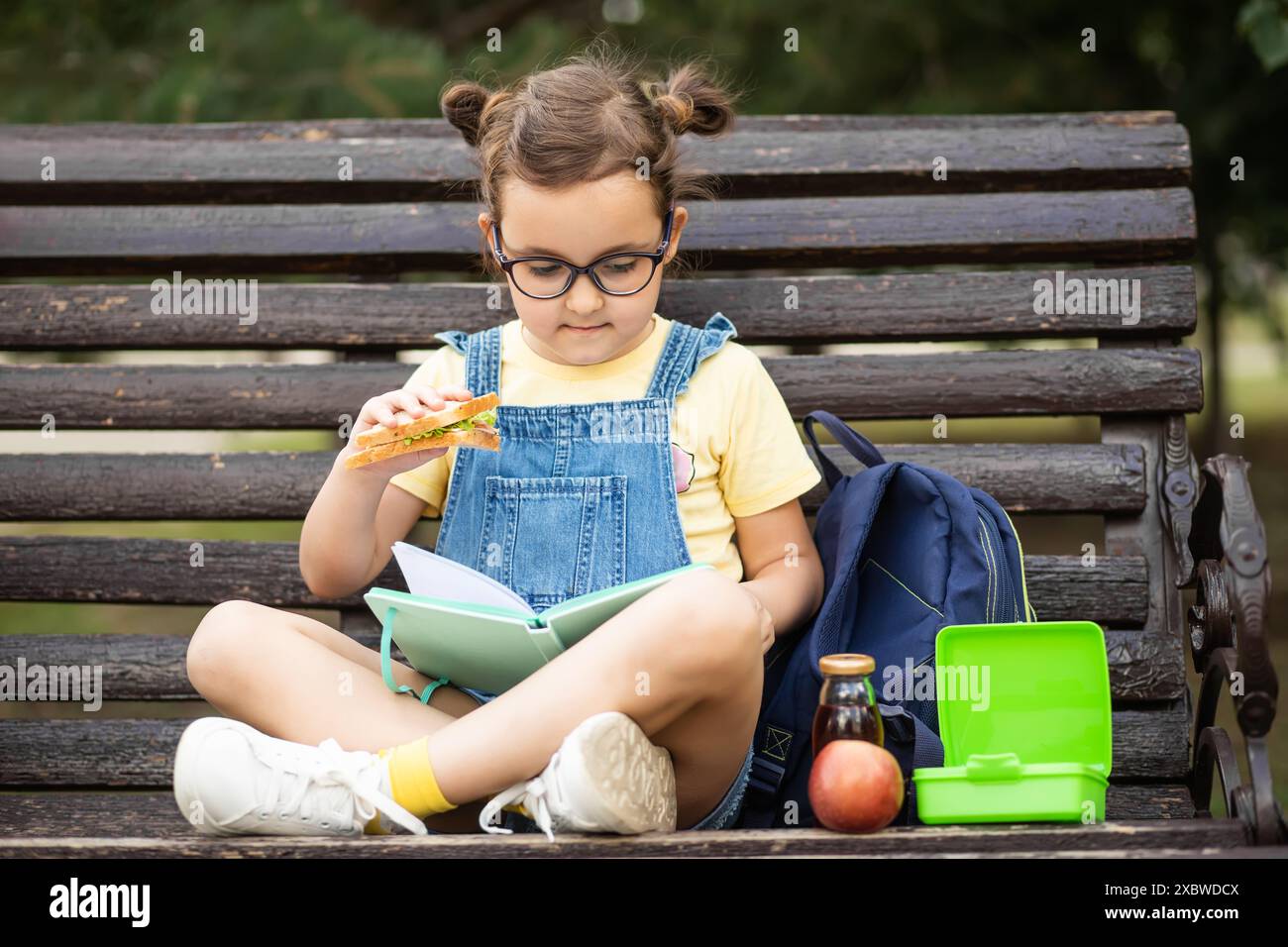 Cute little schoolgirl eating from lunch box outdoor sitting on a bench ...