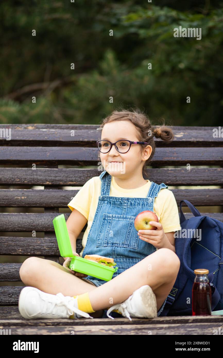 Cute little schoolgirl eating from lunch box outdoor sitting on a bench ...