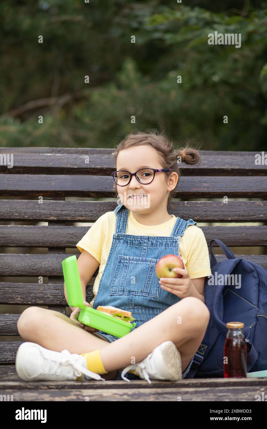 Cute little schoolgirl eating from lunch box outdoor sitting on a bench ...