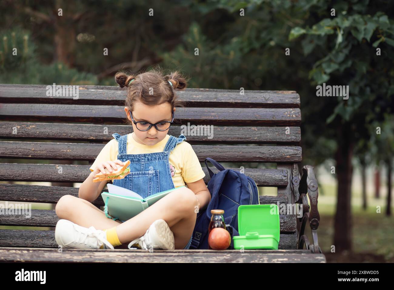 Cute little schoolgirl eating from lunch box outdoor sitting on a bench ...