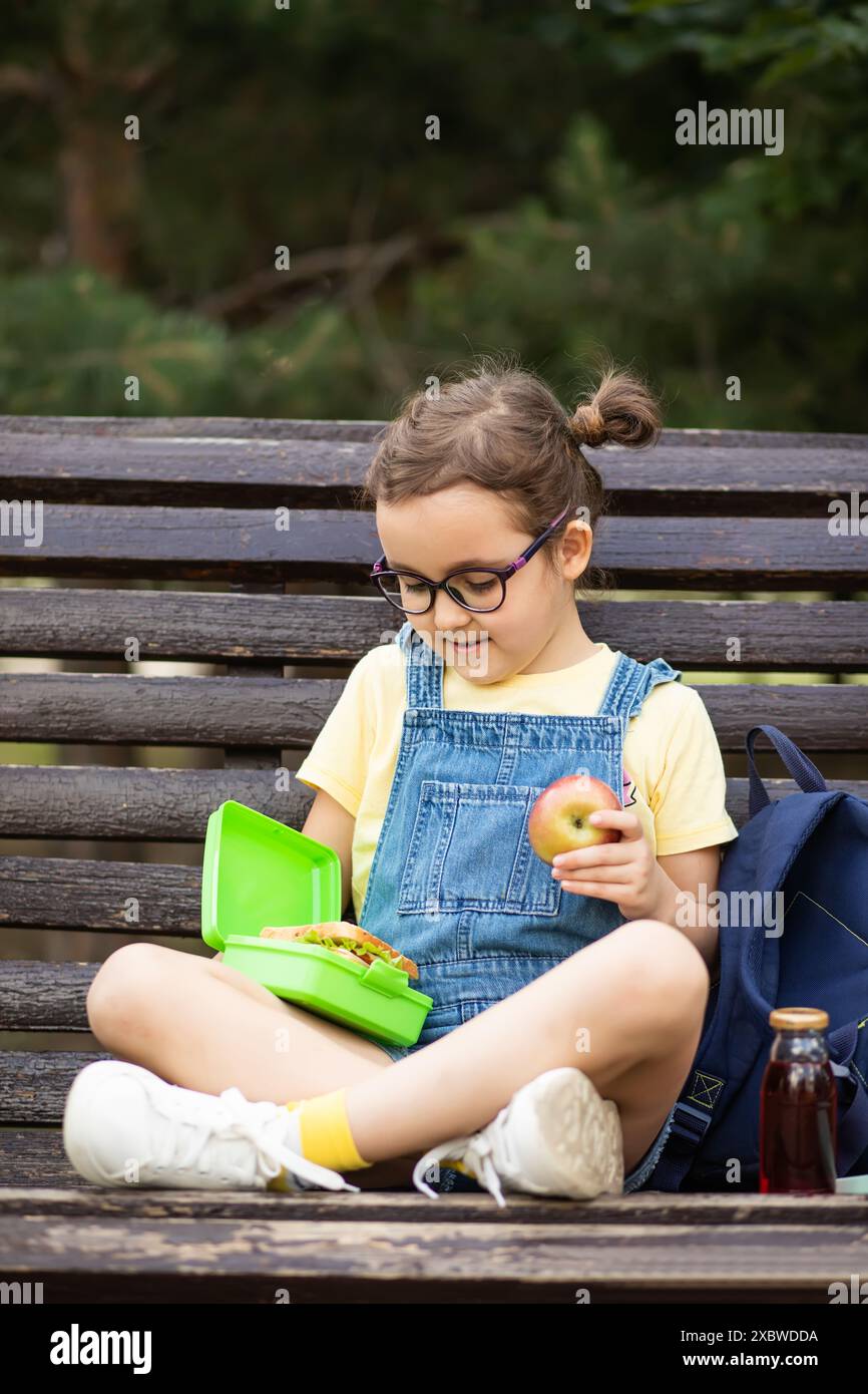 Cute little schoolgirl eating from lunch box outdoor sitting on a bench ...