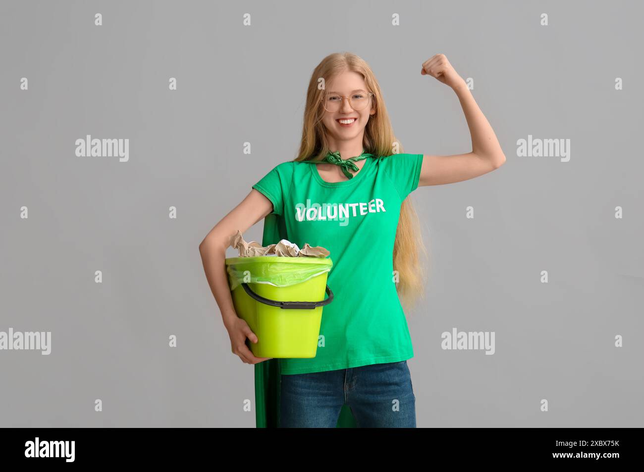 Female volunteer in superhero cape holding recycle bin with paper and showing muscles on grey ...