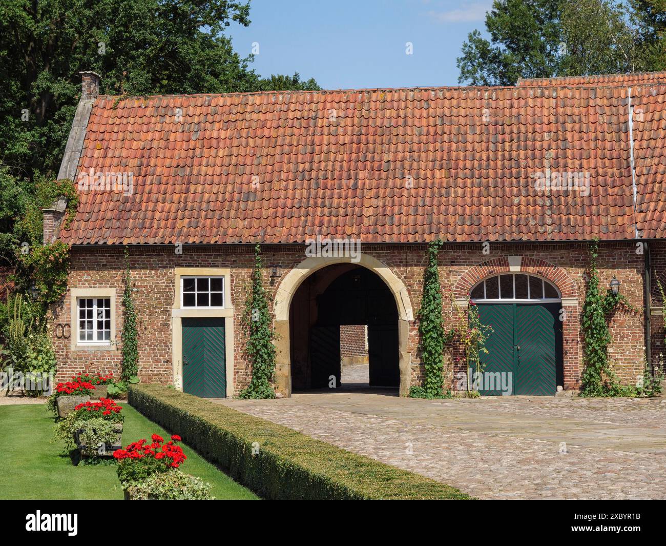 A brick barn with a red roof and courtyard gate, surrounded by a paved ...