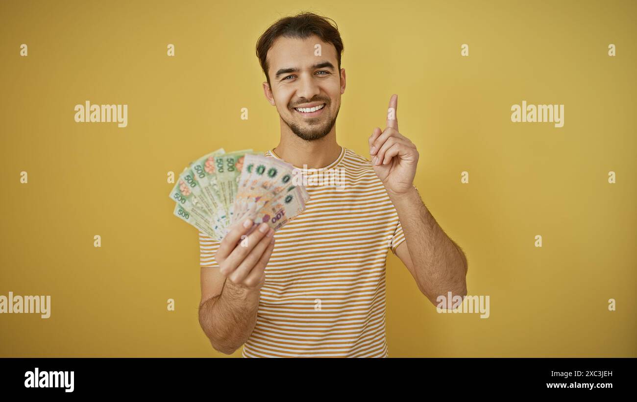 Smiling young man holding argentine pesos against a yellow isolated ...