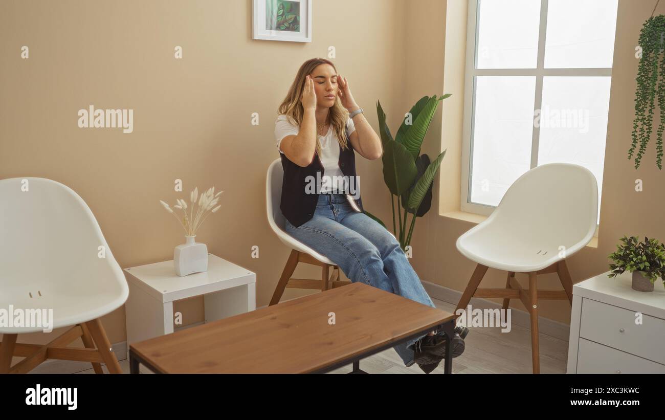 A young blonde woman sits in a waiting room, indoors, looking stressed ...