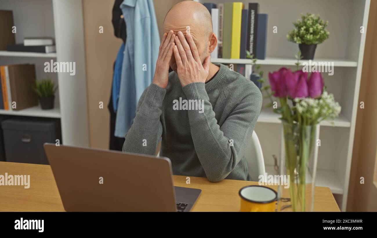 A distressed bald man indoors covering face with hands in a home office ...