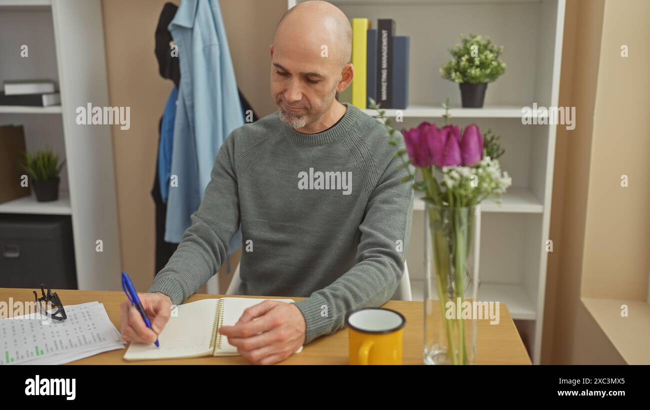 Bald, bearded man writing in a notebook at home, with flowers and ...