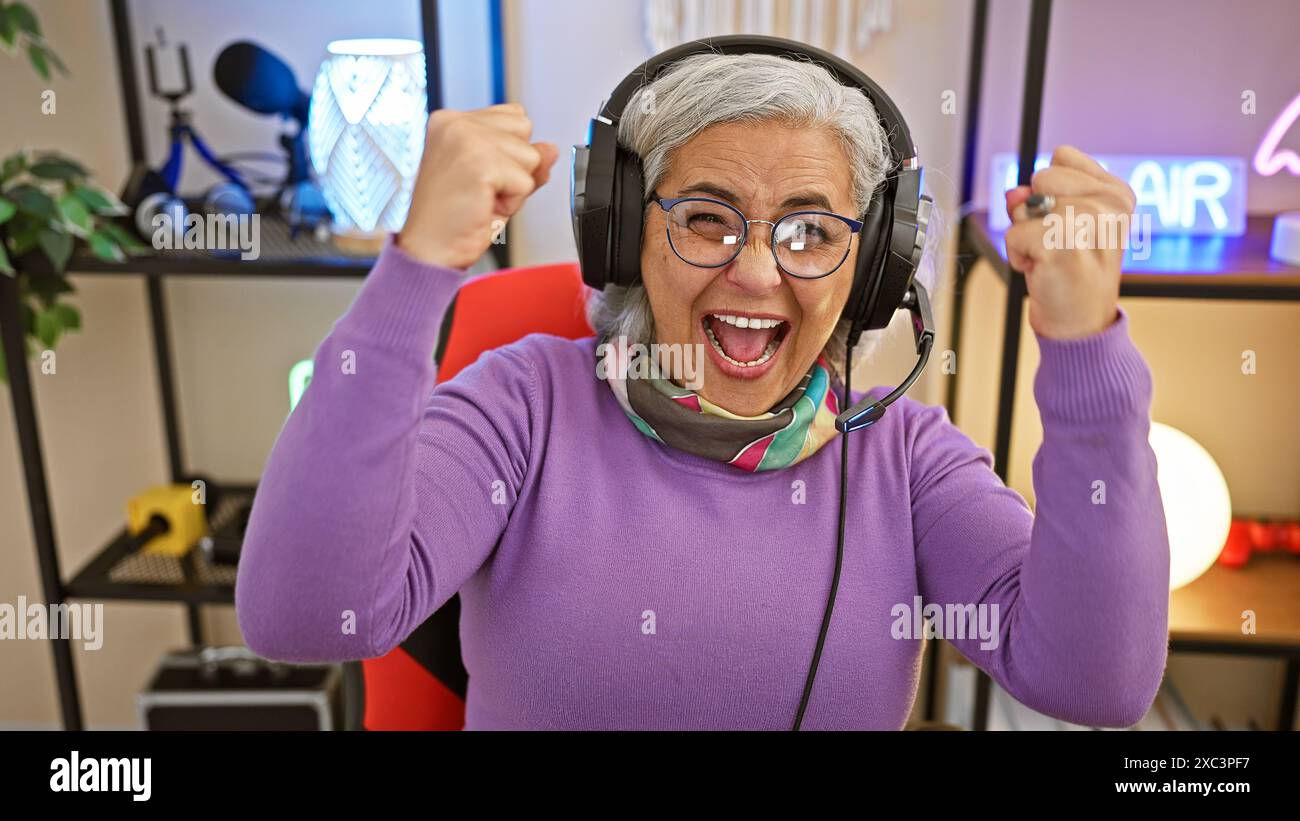 Excited senior woman with headphones enjoys gaming in a well-lit room ...