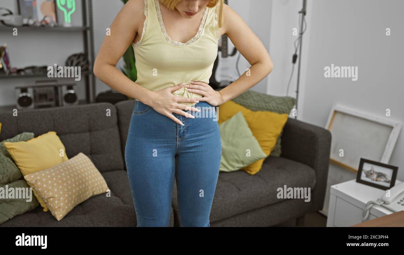 Blonde woman in casual attire standing indoors experiencing stomach ...