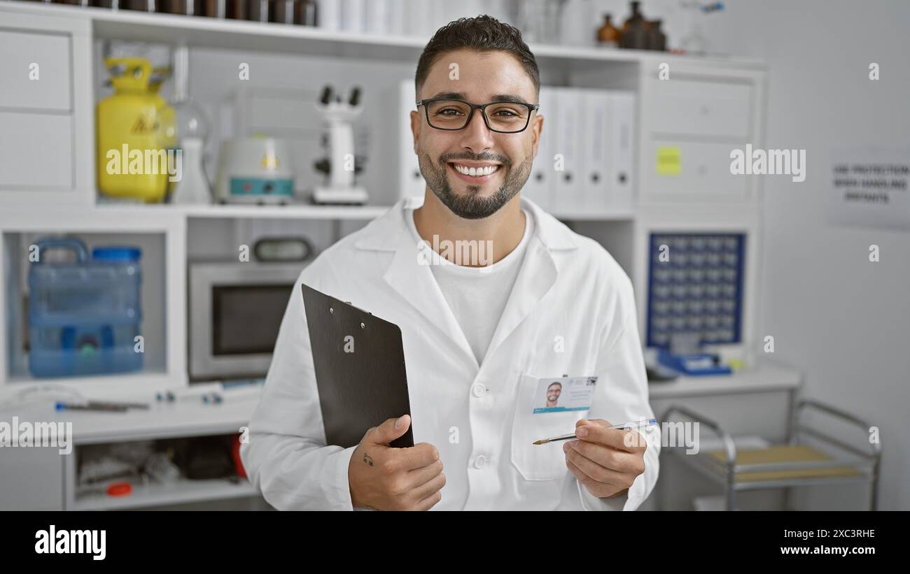 Smiling man with beard wearing glasses posing in a lab coat holding a clipboard and id card in a ...