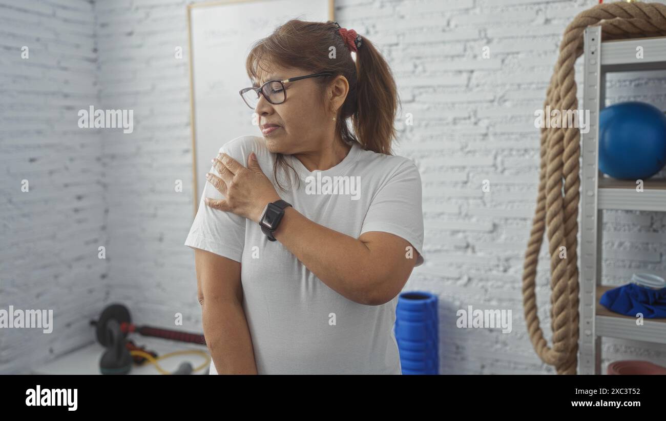 A middle-aged hispanic woman in a gymnasium touches her shoulder in ...