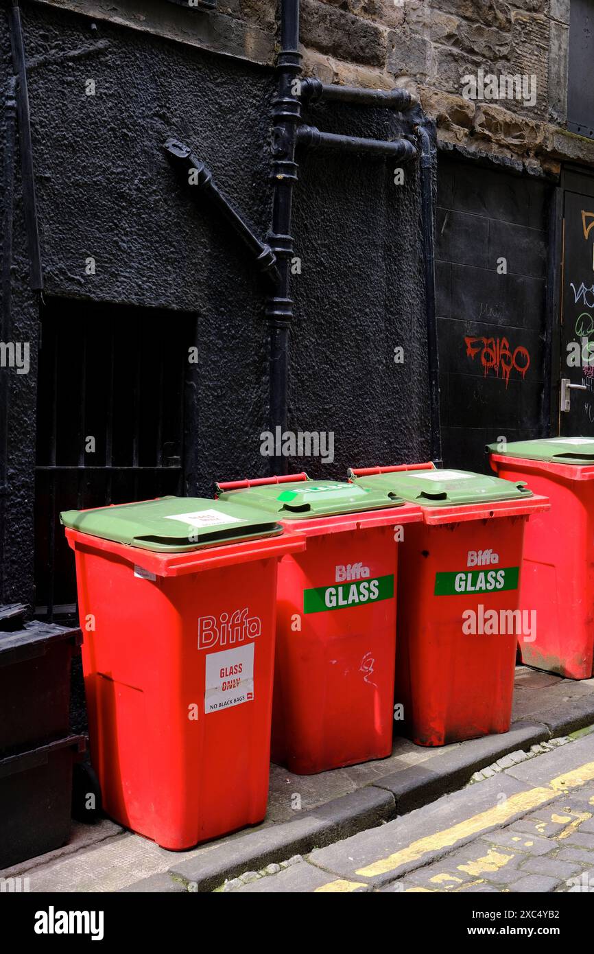 Red commercial glass recycling bins with green lid lined up in front of a black wall outside of ...