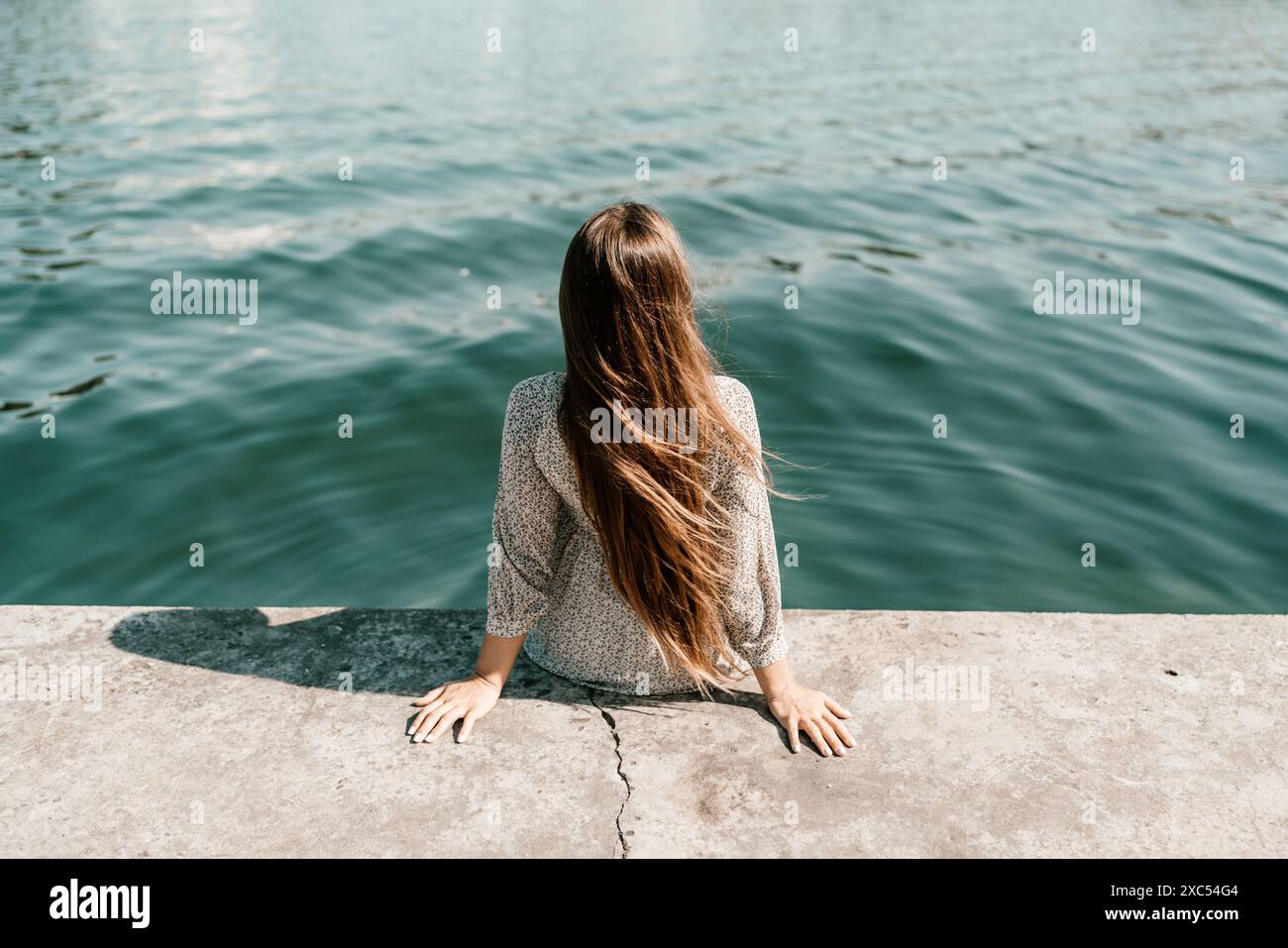 A woman is sitting on a ledge by a body of water, wearing a dress with ...