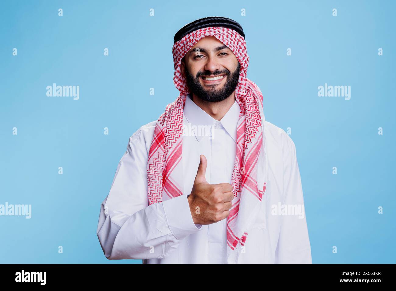Smiling muslim man in white thobe and checkered headdress showing thumb ...