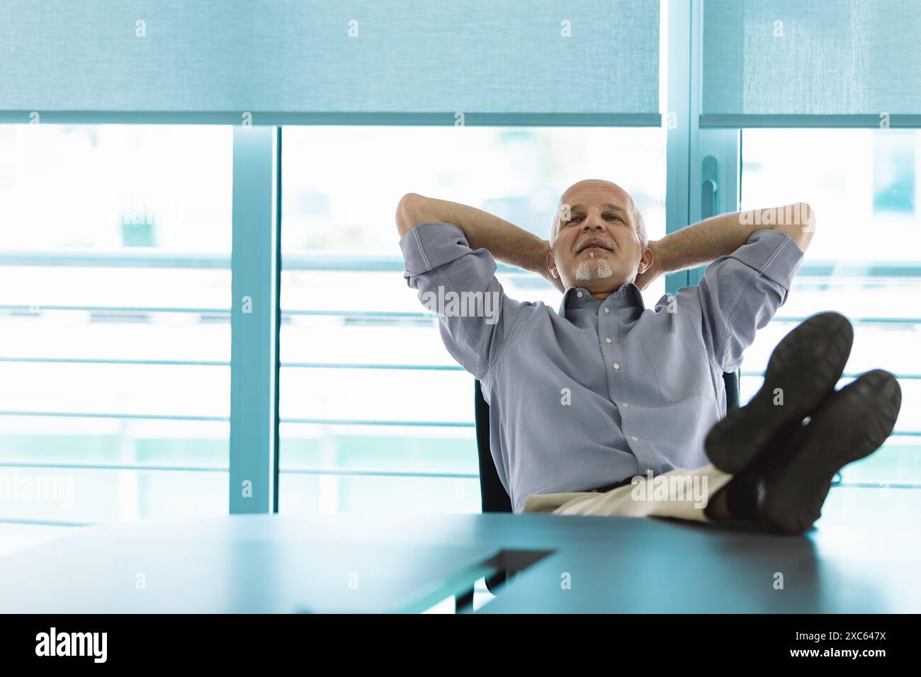 Bald man with white goatee and light blue shirt sits back in a chair ...