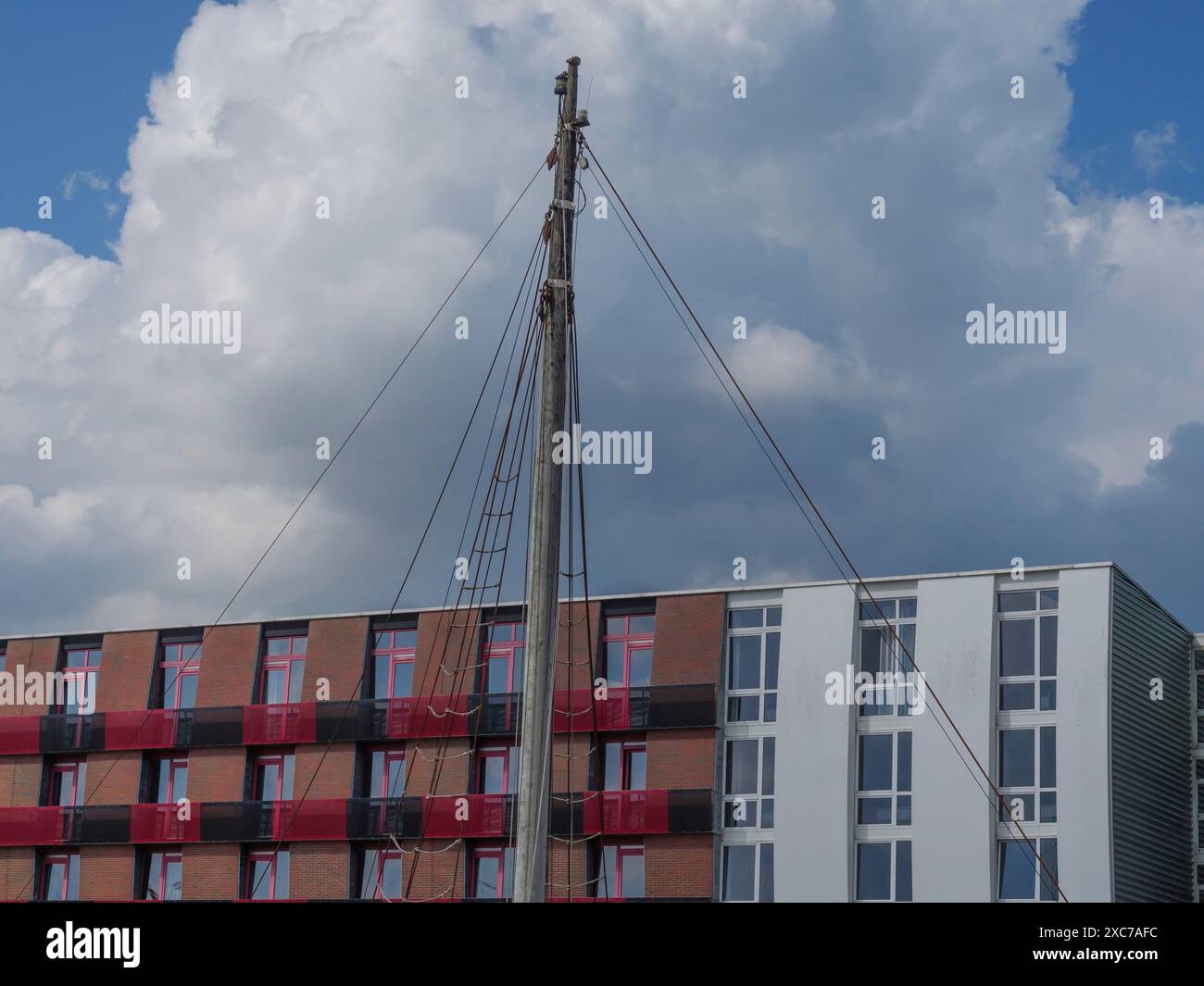 A mast of a sailing boat rises into a cloudy sky in front of a modern ...