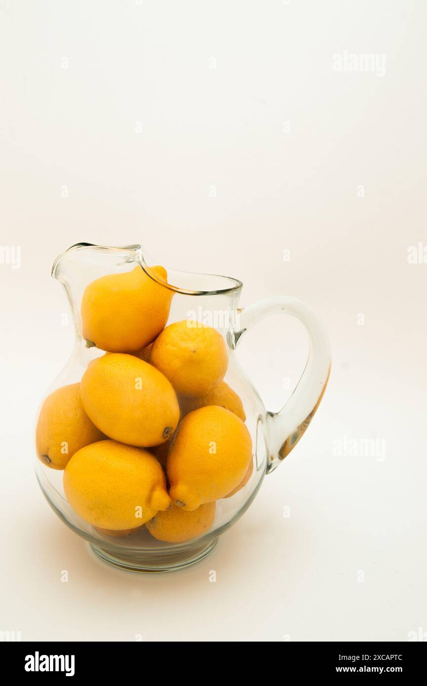 A clear glass pitcher full of whole fresh lemons on a white background ...