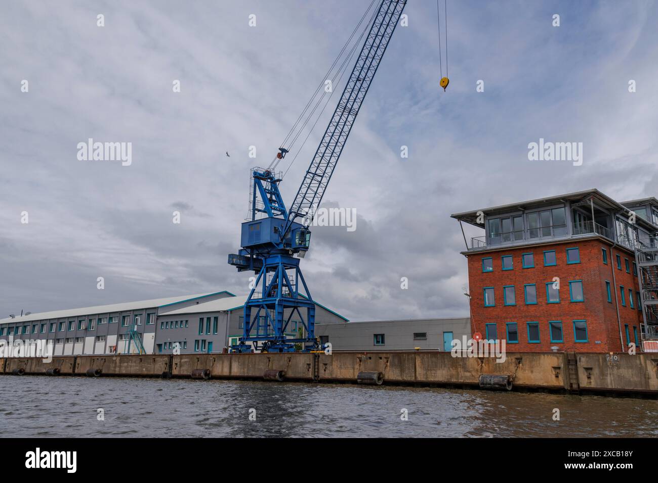 A blue harbour crane next to a brick building against a grey, cloudy ...